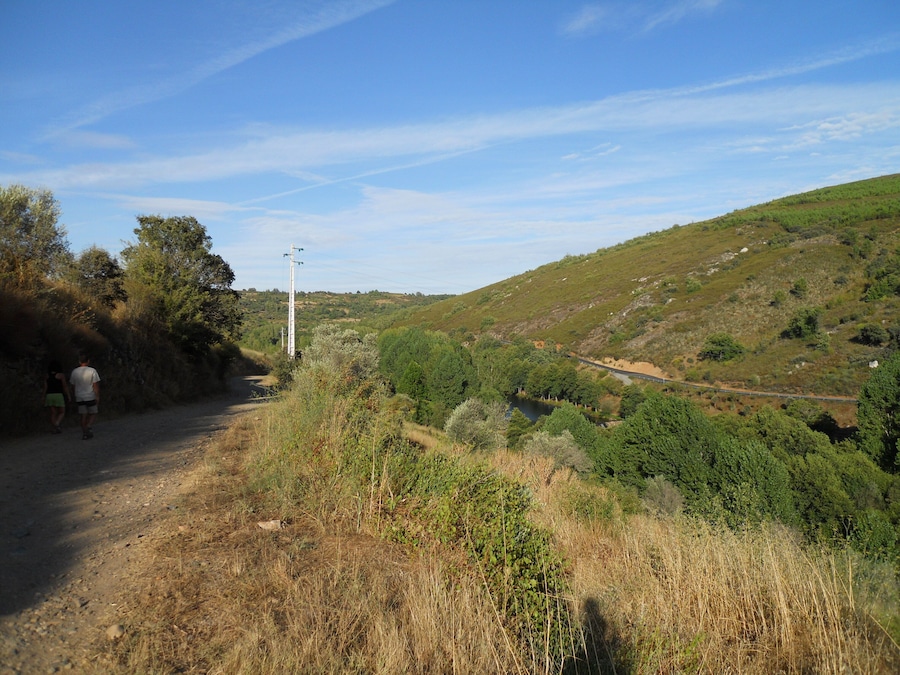 Uma estrada de terra com caminhantes, uma linha de energia e uma paisagem acidentada com vegetação escassa.
