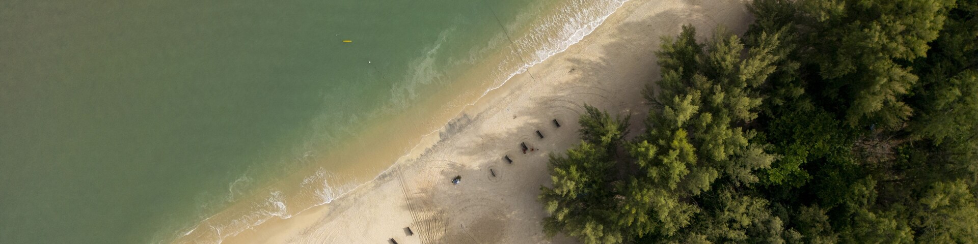 Vista aérea de uma praia com uma fileira de cabanas de praia, uma pequena construção e uma área florestal densa.