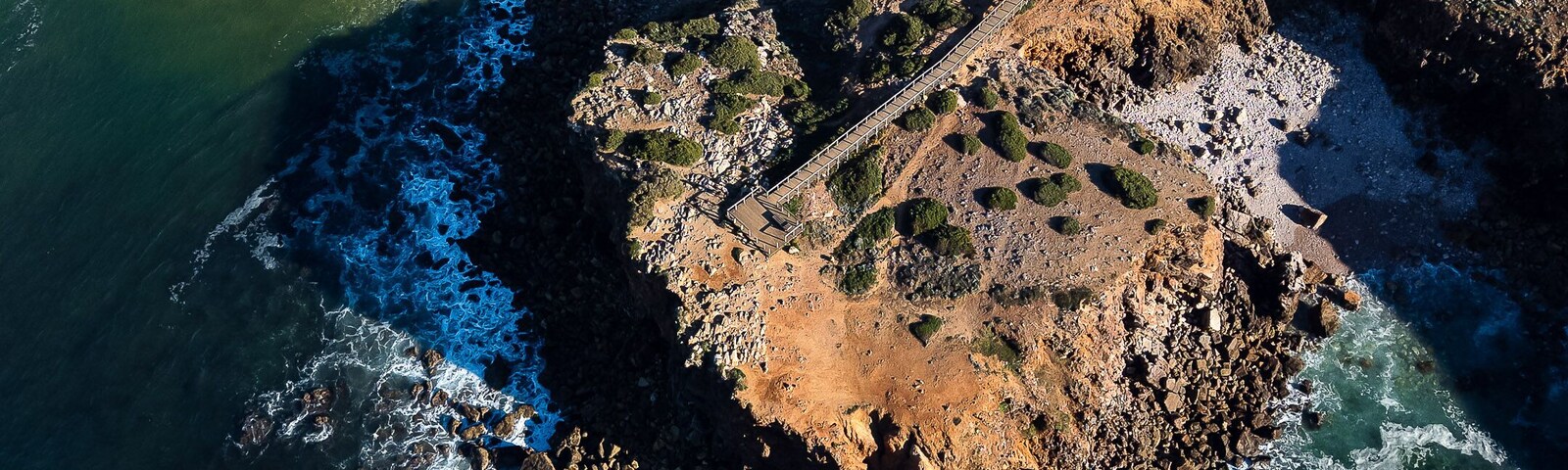 Vista aérea de uma área costeira com praia de areia, falésias rochosas e um caminho à beira-mar.