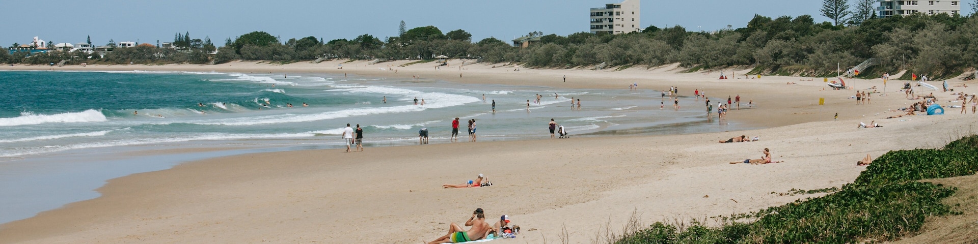 Uma praia arenosa com pessoas tomando sol e nadando, céu azul claro e edifícios ao fundo.