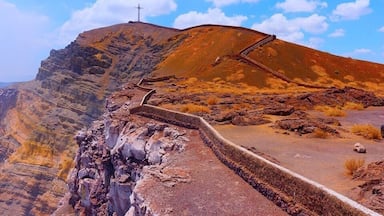 Montanha rochosa e íngreme com uma cruz no topo e uma estrada sinuosa descendo.