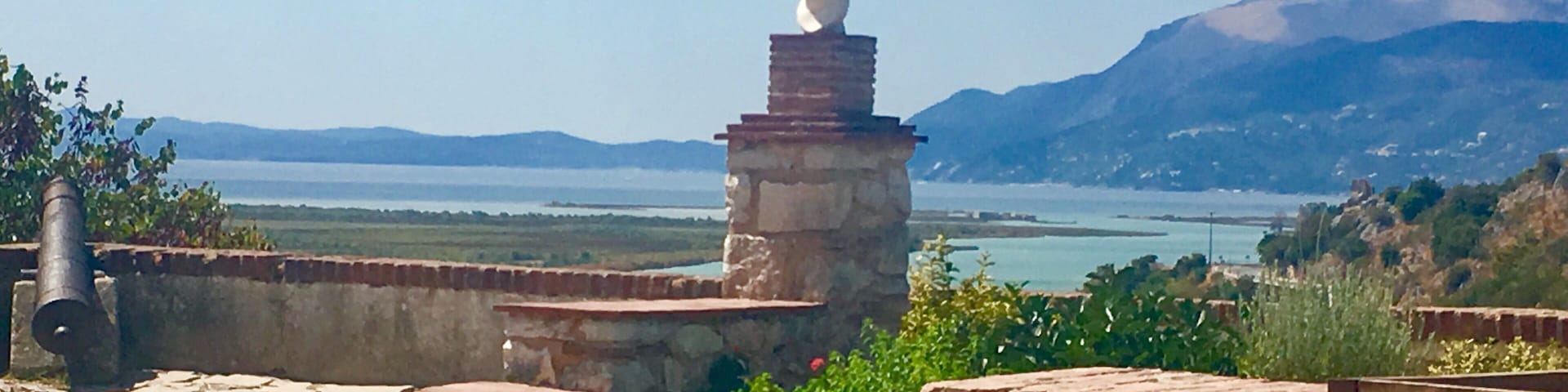 Uma fortaleza costeira com uma estátua, paredes de pedra e vista para o mar e montanhas.