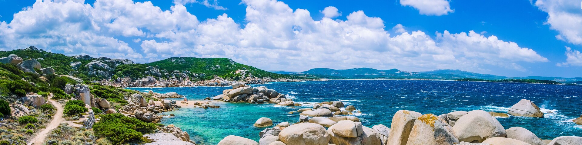 Paisagem costeira com grandes pedras, mar azul límpido e colinas verdes.