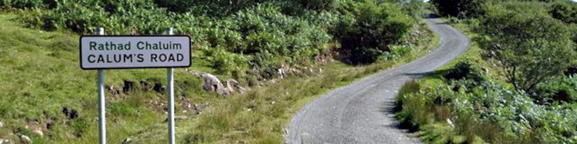 Uma estrada sinuosa chamada Rathad Chaluim CALUM'S ROAD, cercada por vegetação exuberante e terreno rochoso.