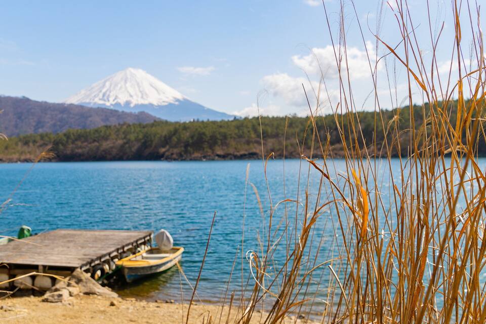 Um lago com um barco atracado na margem, uma montanha ao fundo e gramíneas altas em primeiro plano.