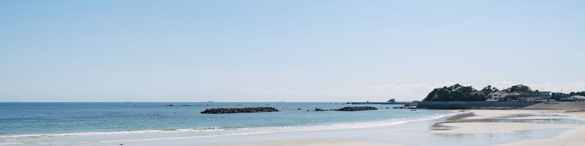 Uma praia com uma pessoa carregando uma prancha de surf, águas calmas e um afloramento rochoso ao longe.
