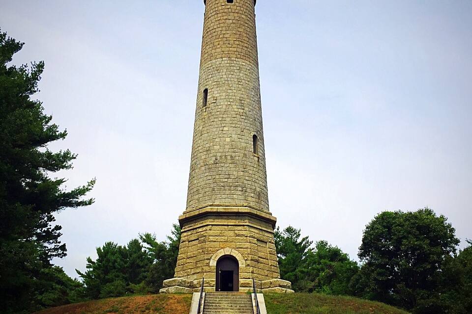 Uma torre de pedra alta com uma estátua no topo, cercada por árvores e terreno gramado.