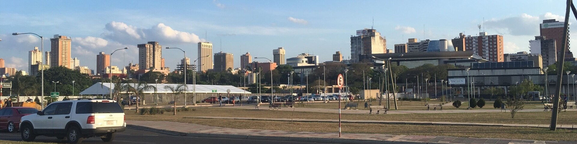 Paisagem urbana com edifícios altos, um SUV branco na rua e uma faixa de pedestres.