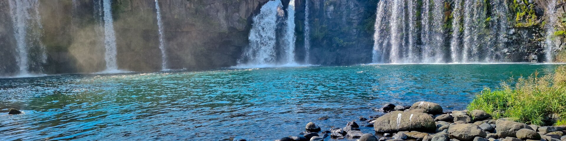 Uma cascata deságua em um lago azul e límpido, rodeado por pedras e vegetação.