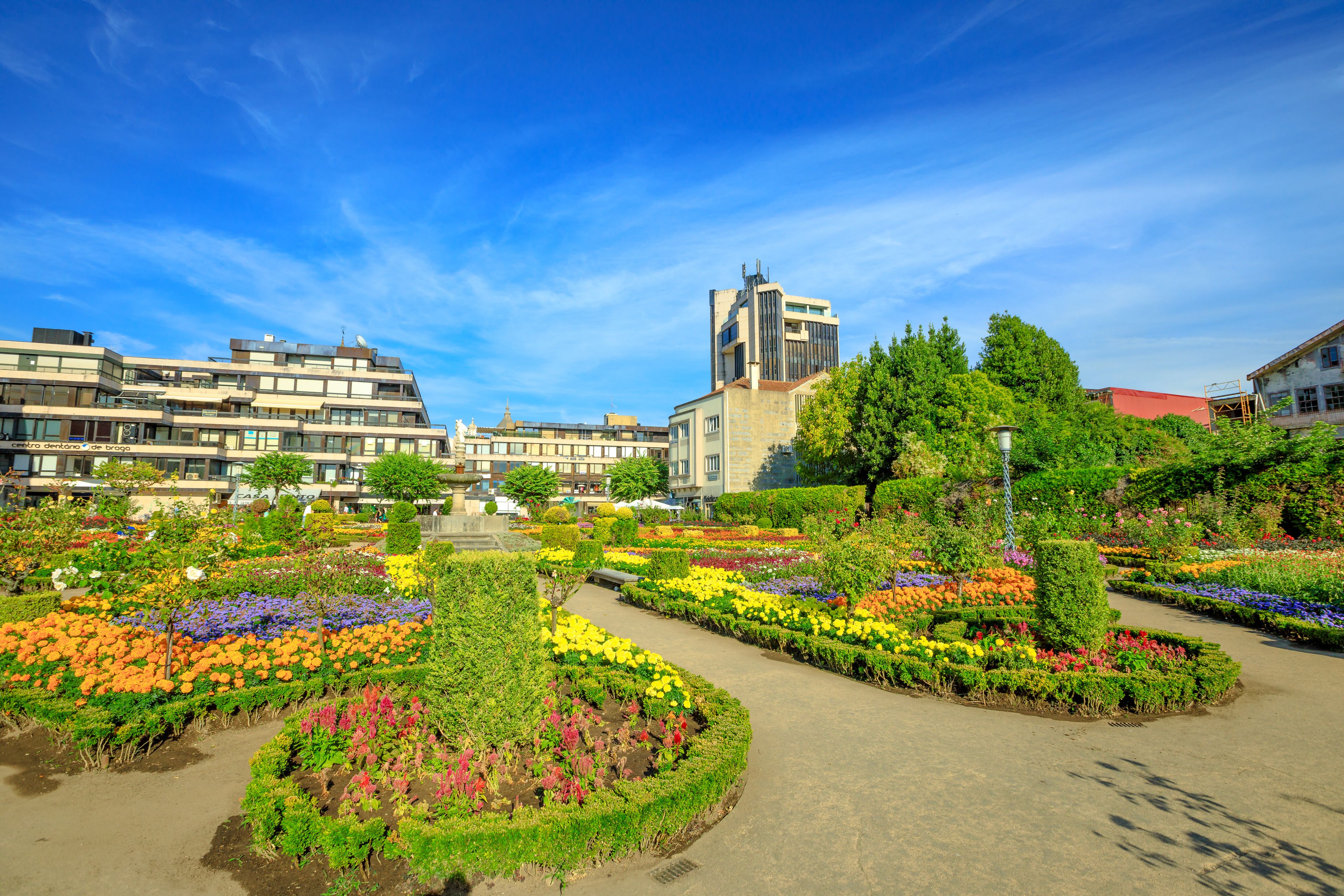 Um jardim florido e vibrante com diversas flores coloridas, um complexo de edifícios modernos e um céu azul claro.