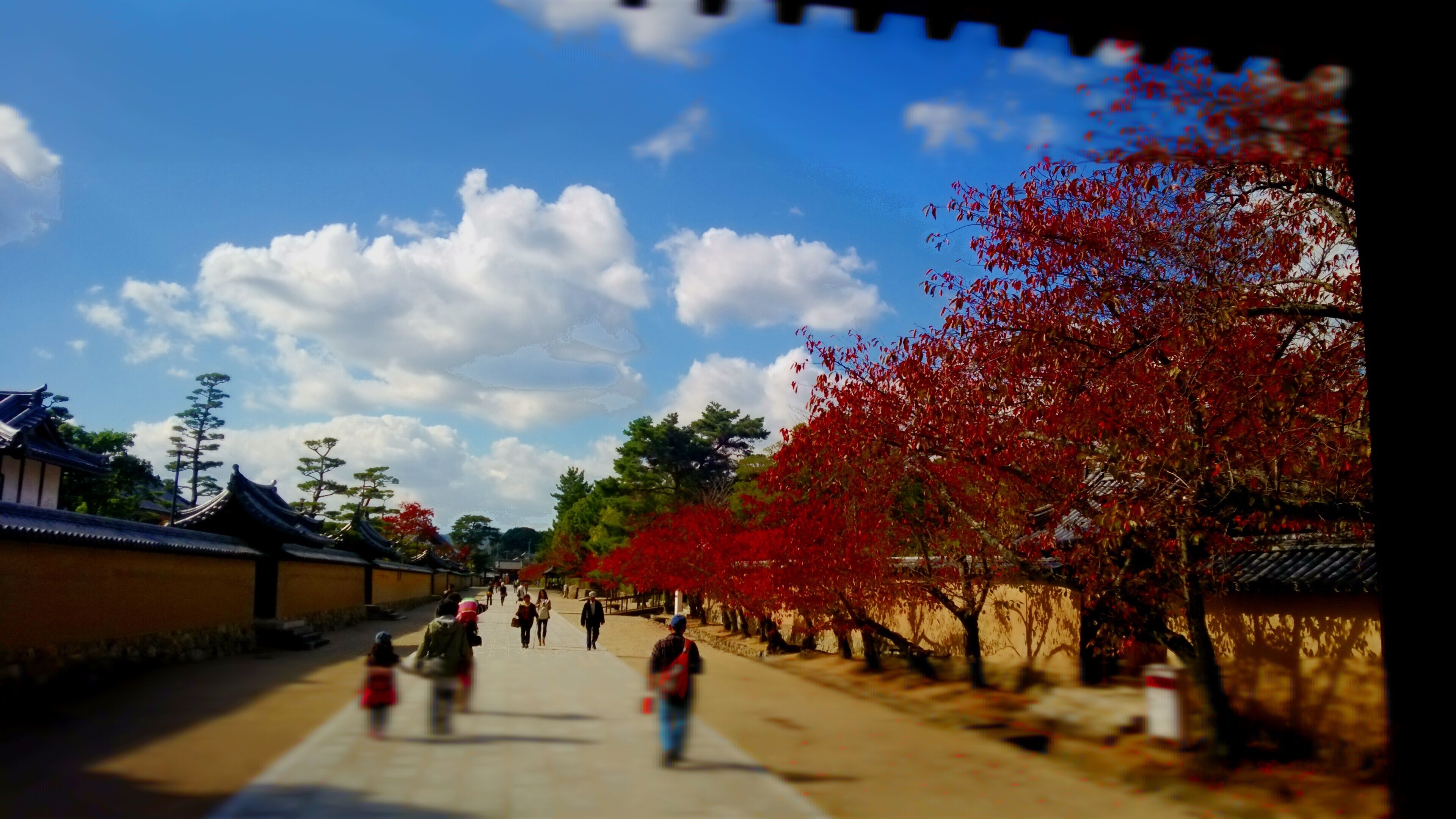 Uma rua tradicional japonesa com folhas avermelhadas de outono, pessoas caminhando e edifícios tradicionais.