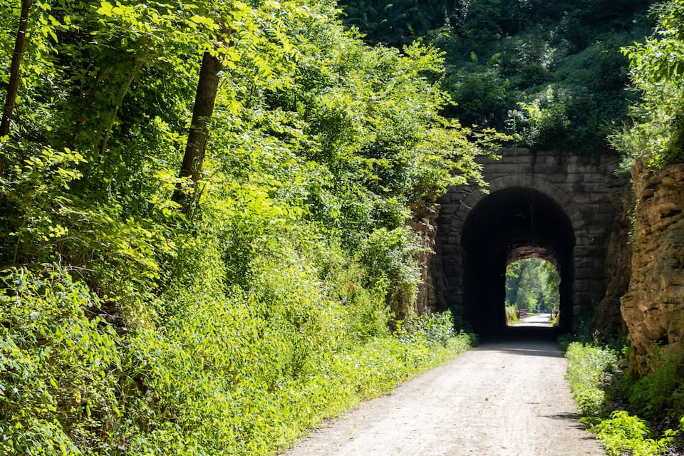 Uma estrada de terra que leva a uma ponte de pedra arqueada, cercada por vegetação exuberante.