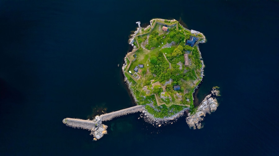 Vista aérea de uma pequena ilha com uma estrutura fortificada, vegetação exuberante e uma ponte que a liga ao continente.