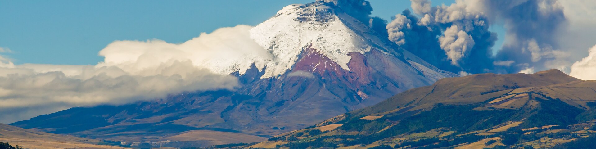 Uma montanha coberta de neve com uma coluna de fumaça, rodeada por colinas e vales ondulantes.