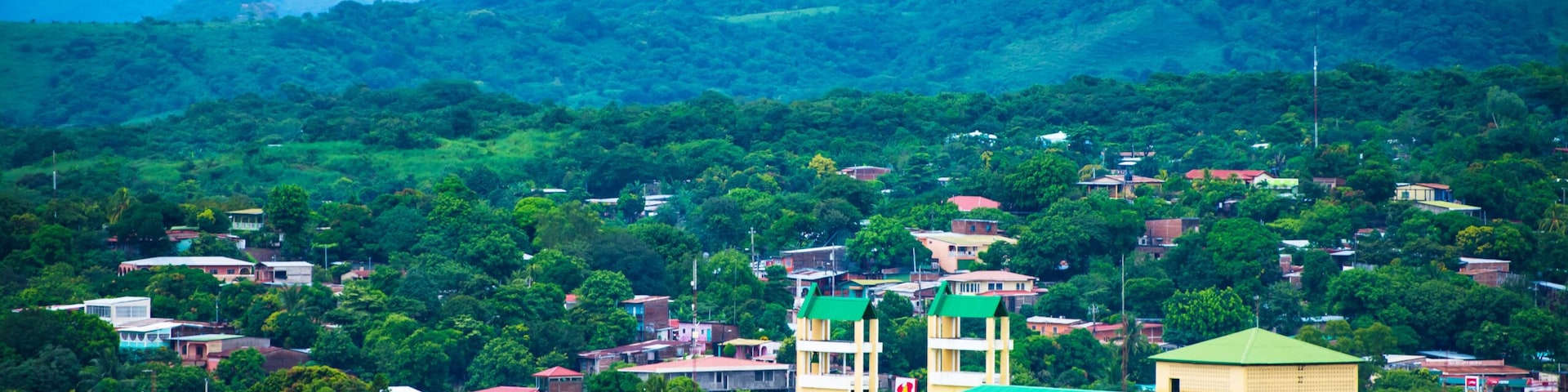 Uma cidade com um edifício amarelo imponente e telhado verde, rodeada por colinas verdejantes e casas dispersas.
