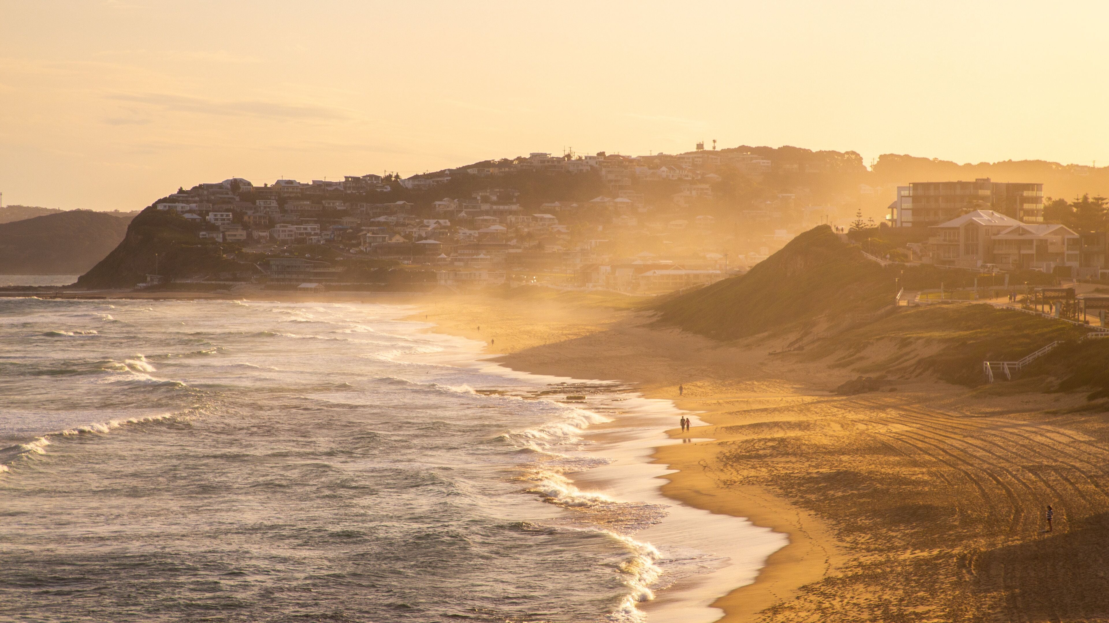 Paisagem costeira com ondas quebrando na praia, uma faixa de areia e uma encosta com edifícios.