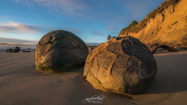 Grandes pedras em uma praia arenosa, com um penhasco ao fundo.