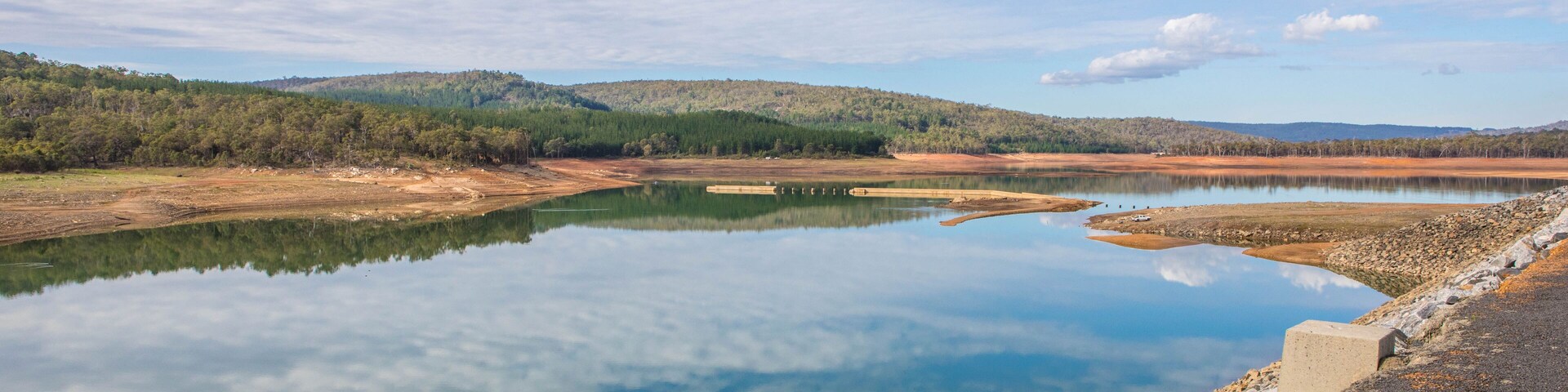 Um lago calmo com margem rochosa, rodeado por colinas arborizadas sob um céu parcialmente nublado.