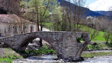 Uma ponte de pedra sobre um rio, rodeada por árvores e uma montanha ao fundo.