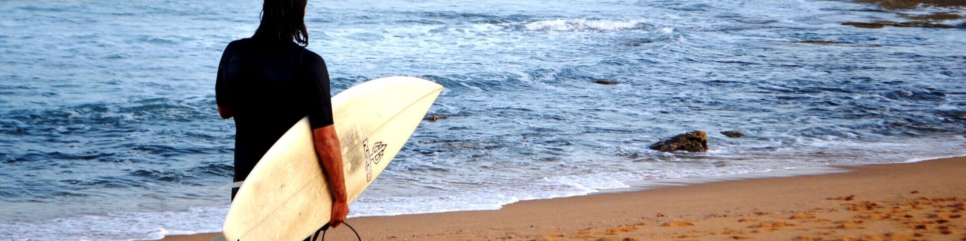 Uma pessoa segurando uma prancha de surf numa praia arenosa, com ondas ao fundo.