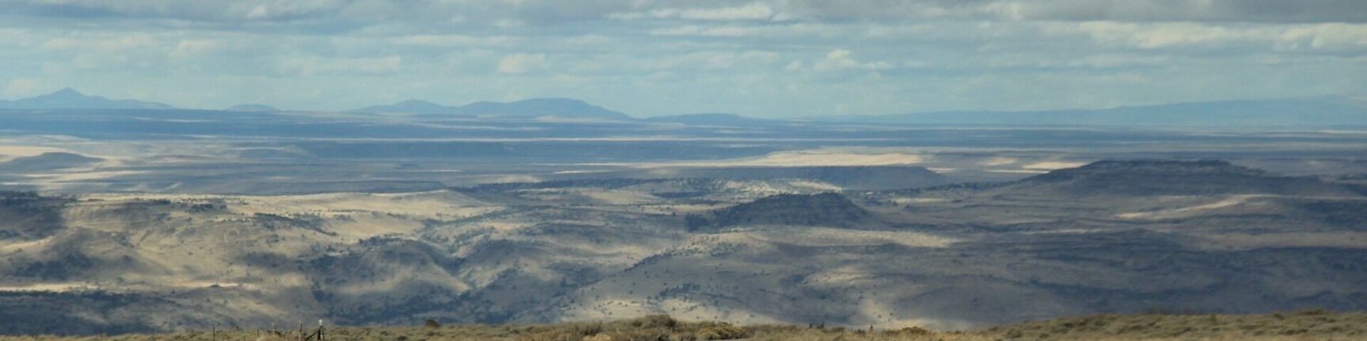 Uma estrada de terra sinuosa atravessa uma paisagem seca e montanhosa, com montanhas distantes sob um céu nublado.