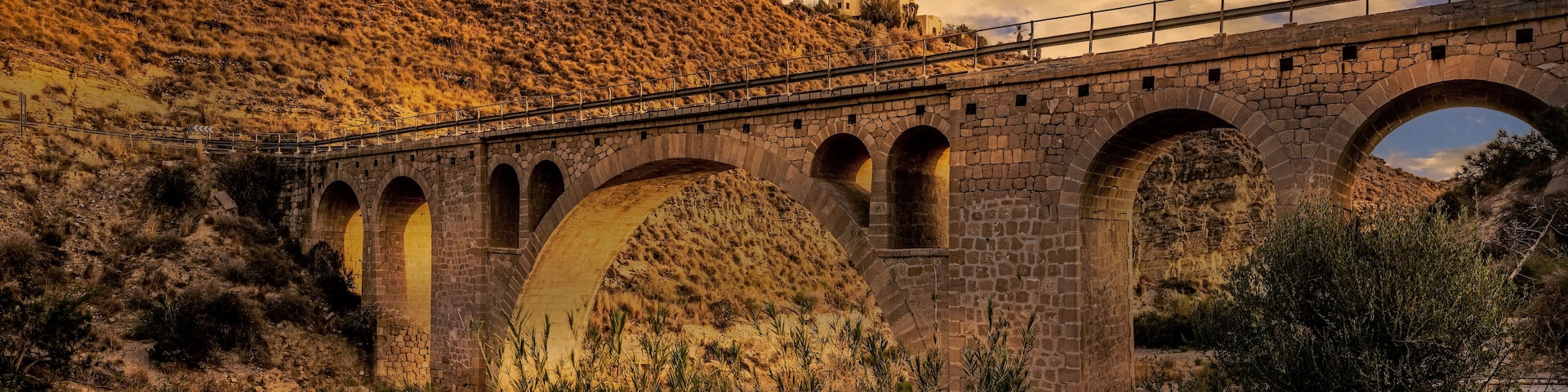 Uma ponte de pedra com várias arcos atravessa um vale, cercada por vegetação seca e uma colina ao fundo.