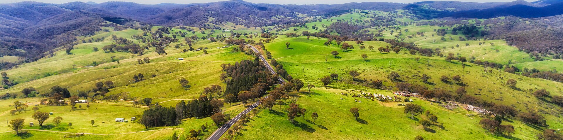 Uma estrada sinuosa serpenteando por colinas verdes ondulantes e montanhas ao longe, sob um céu parcialmente nublado.