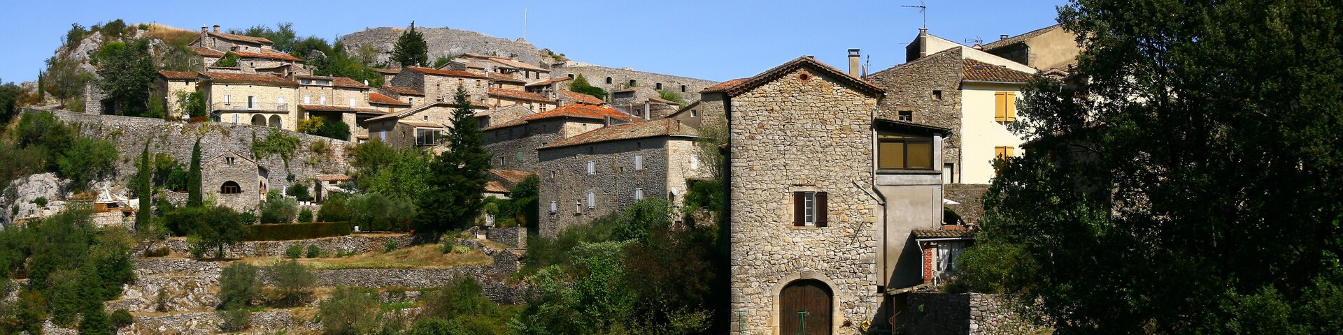 Uma aldeia na encosta de uma colina, com edifícios de pedra, uma igreja e vegetação exuberante.