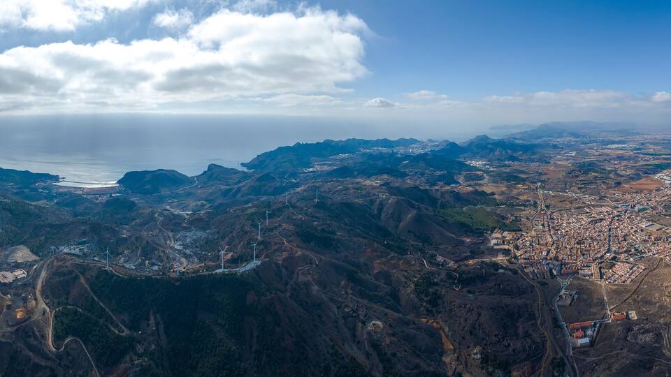 Paisagem montanhosa com uma cidade costeira, turbinas eólicas e uma estrada sinuosa.