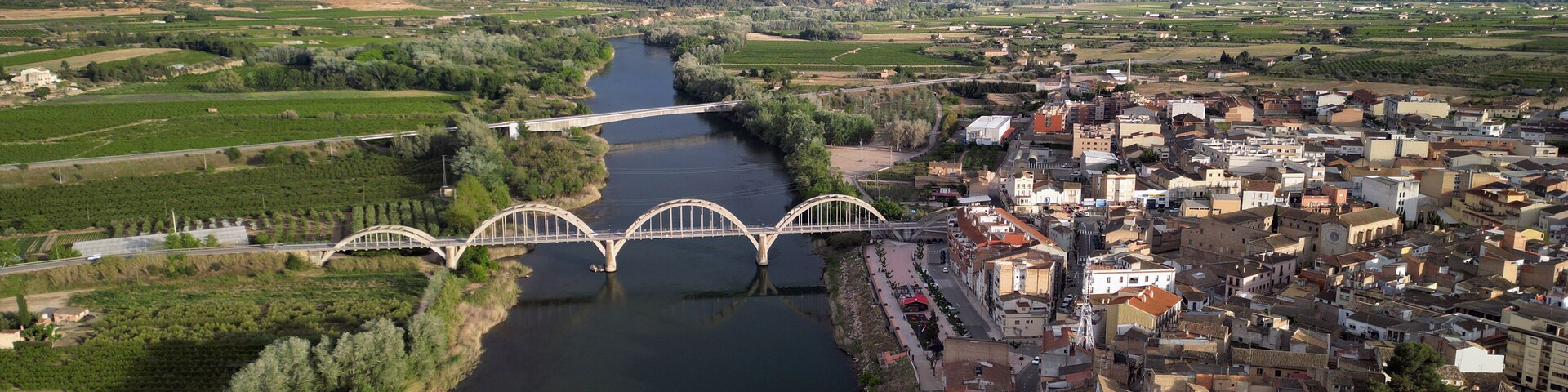 Um rio serpenteia por uma cidade com uma ponte, cercada por montanhas e campos.