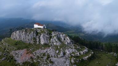 Uma igreja branca numa colina rochosa com telhado vermelho, rodeada por uma paisagem montanhosa e nuvens.