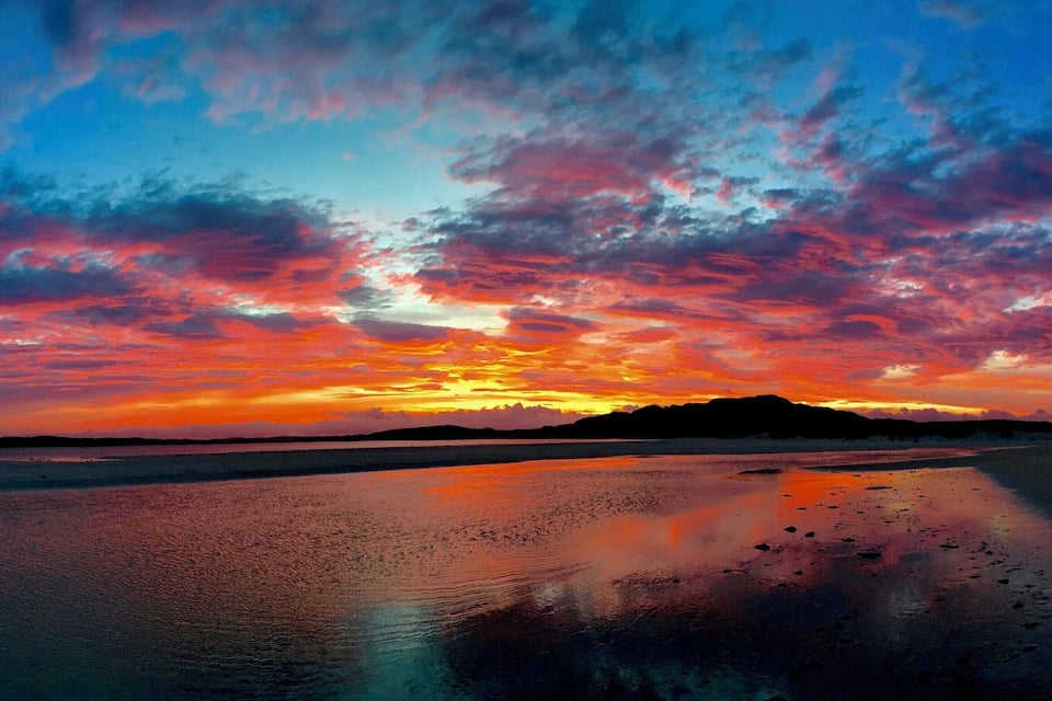 Uma praia ao pôr do sol, com nuvens vibrantes em tons de vermelho e laranja refletindo na areia molhada.