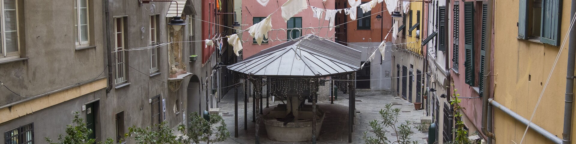Uma rua de paralelepípedos estreita, flanqueada por edifícios coloridos, plantas em vasos e roupas penduradas.