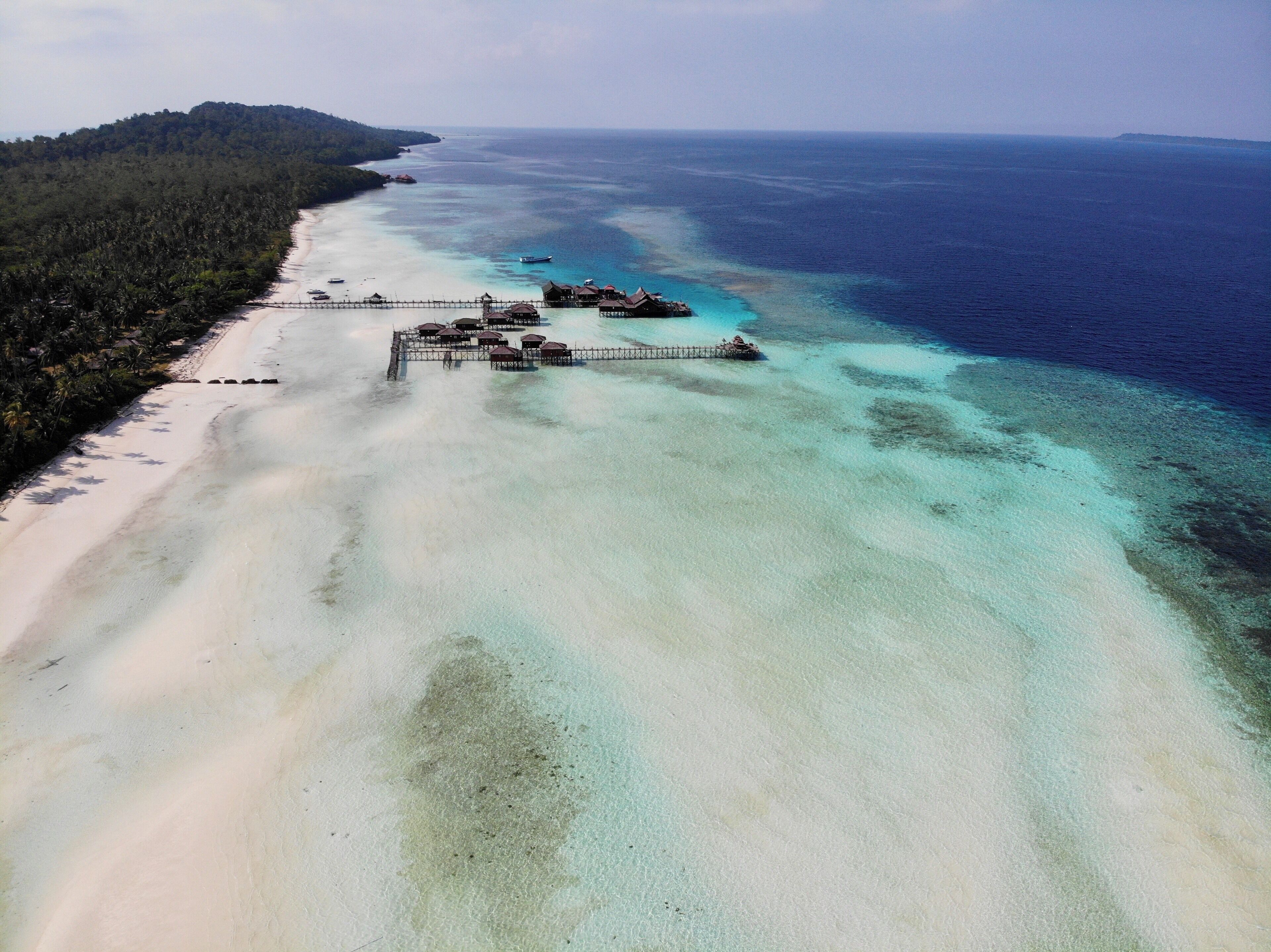 Vista aérea de uma praia com águas azul-turquesa cristalinas, uma faixa de areia e bangalôs sobre a água.