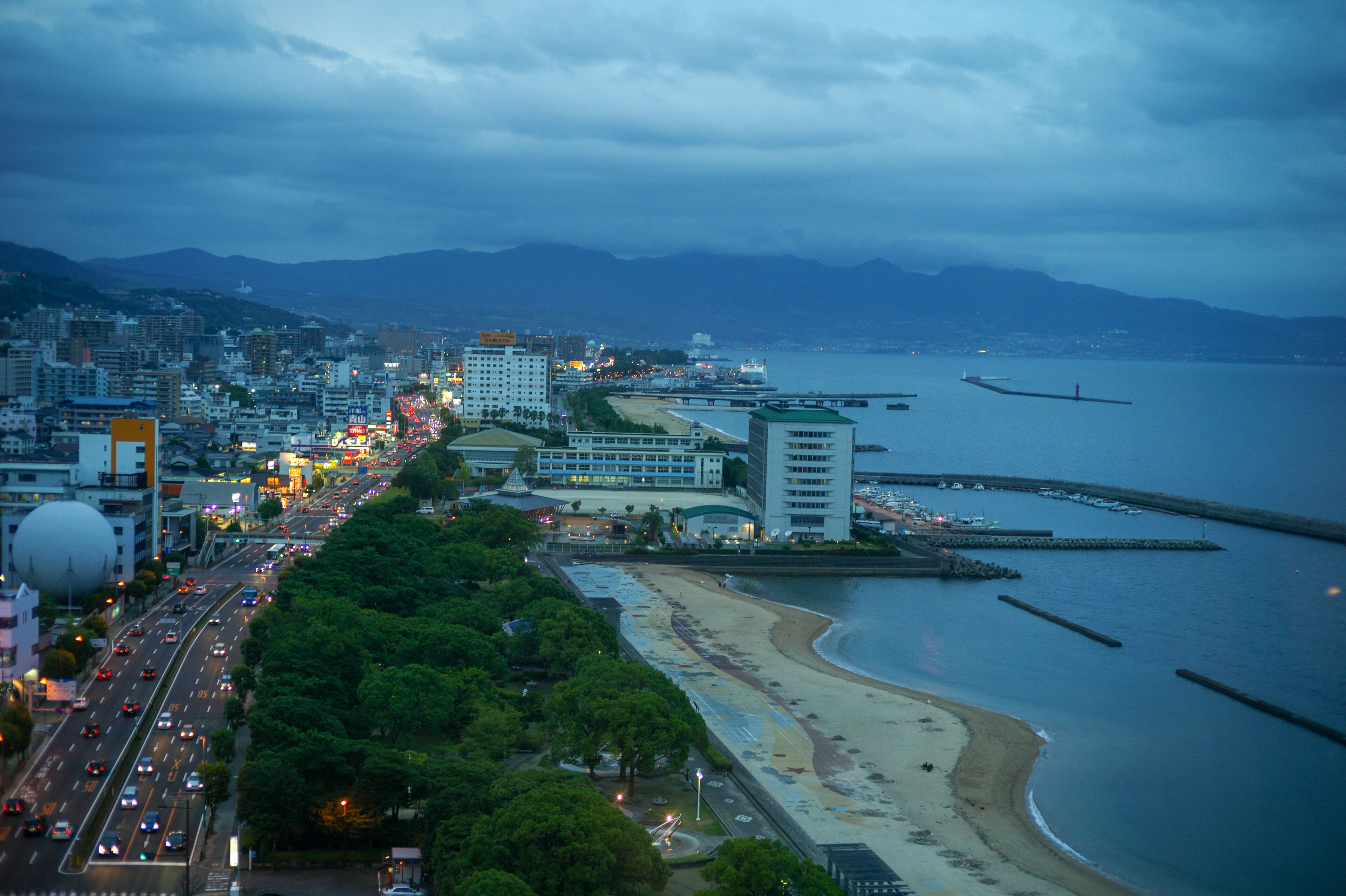 Paisagem costeira com uma rodovia movimentada, uma praia e montanhas ao fundo.