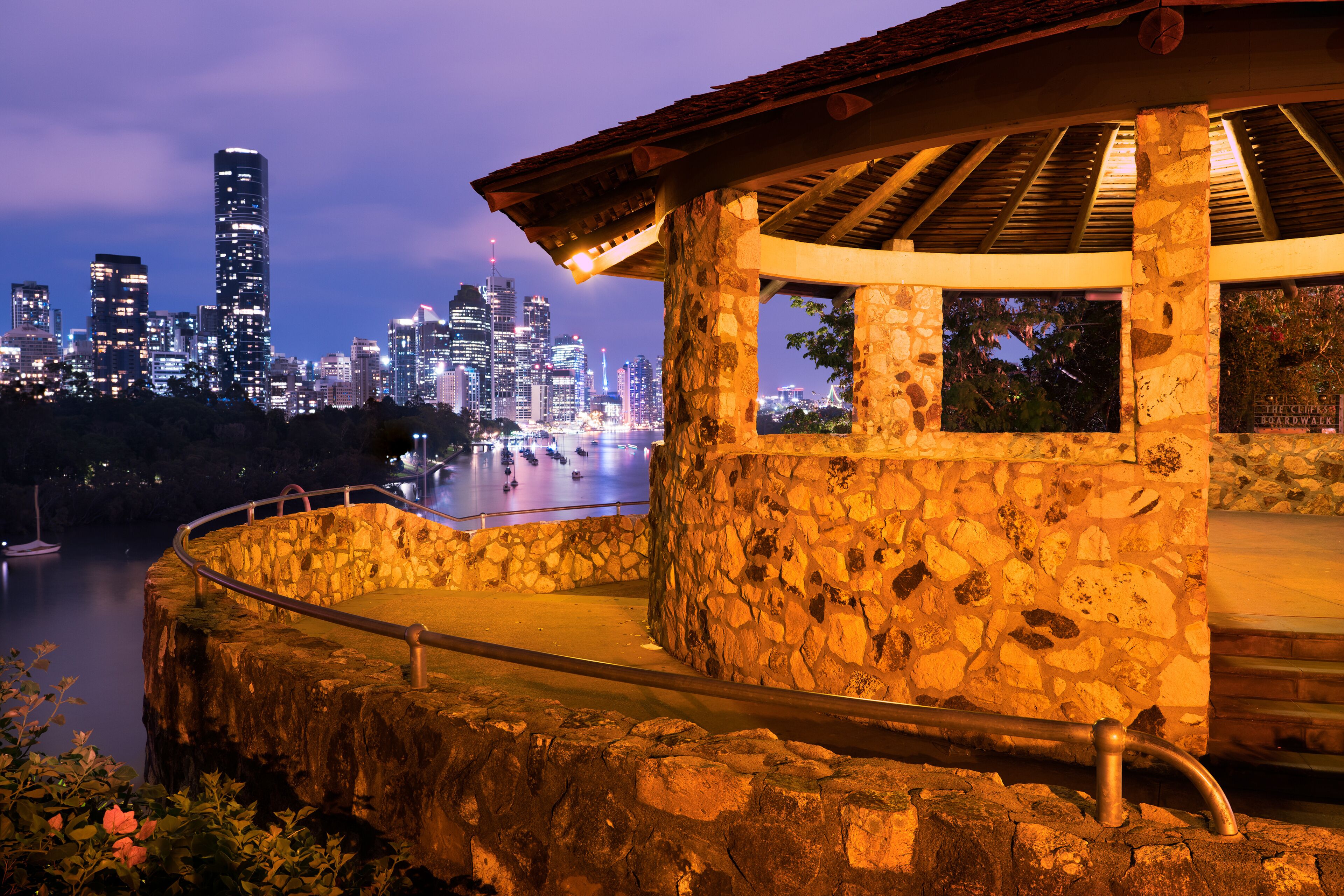 Um pavilhão de pedra com grade de metal, com vista para a orla marítima e horizonte urbano ao fundo.