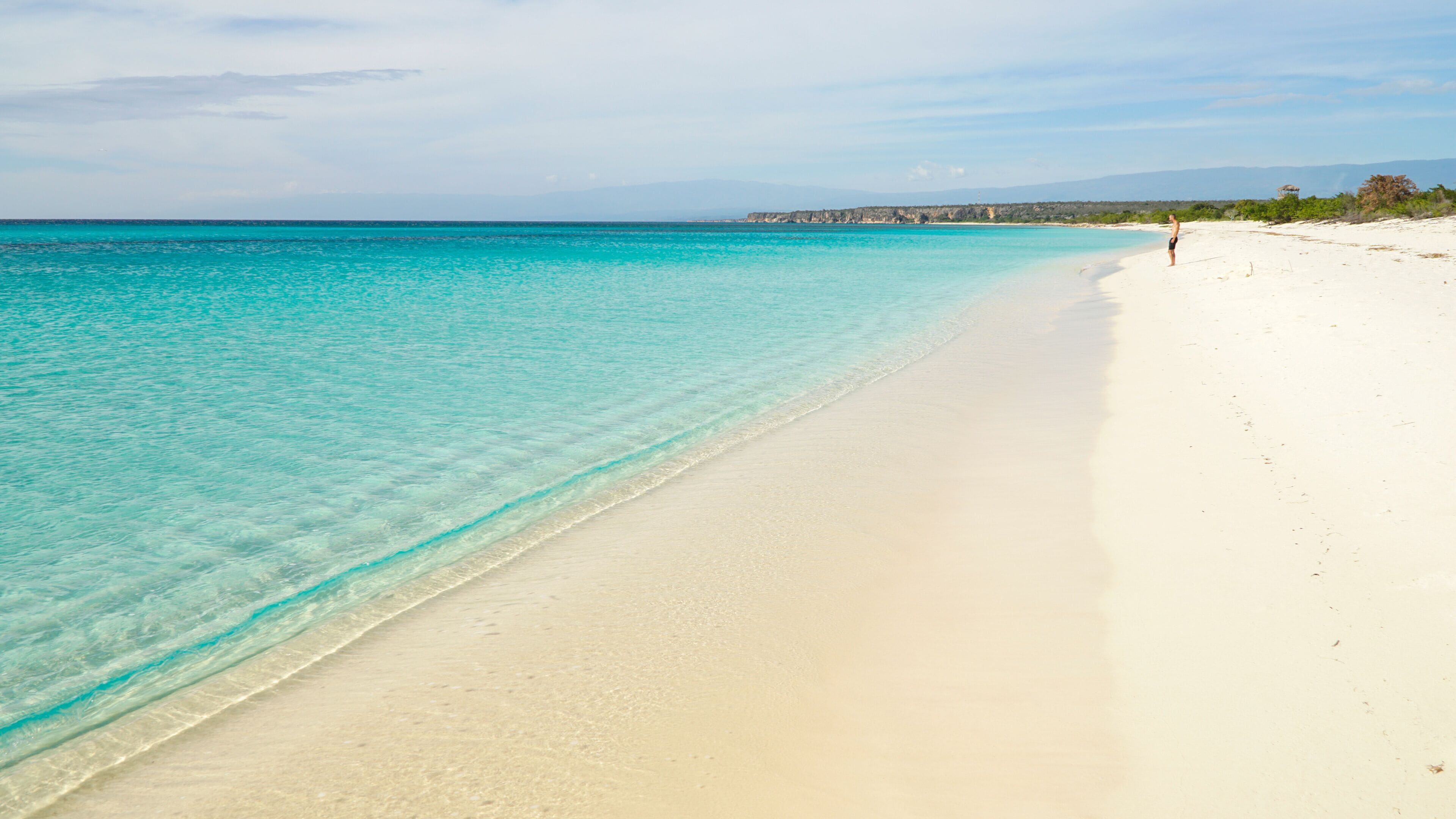 Uma praia com águas turquesa cristalinas, areal arenoso e uma pessoa caminhando pela orla.