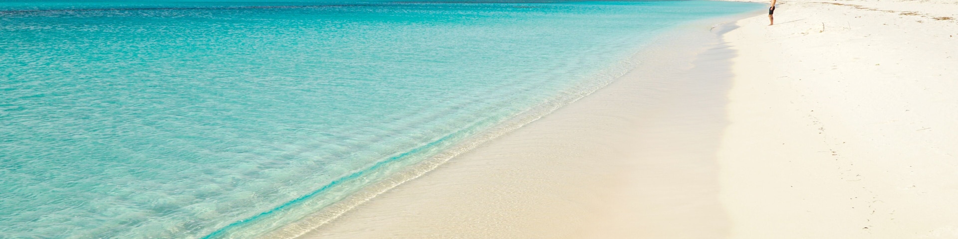 Uma praia com águas turquesa cristalinas, areal arenoso e uma pessoa caminhando pela orla.