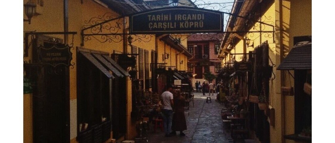 Uma rua estreita com lojas de ambos os lados, uma placa com a inscrição "TARIHI IRGANDI CARSILI KÖPRÜ" no centro e um caminho de calçada.