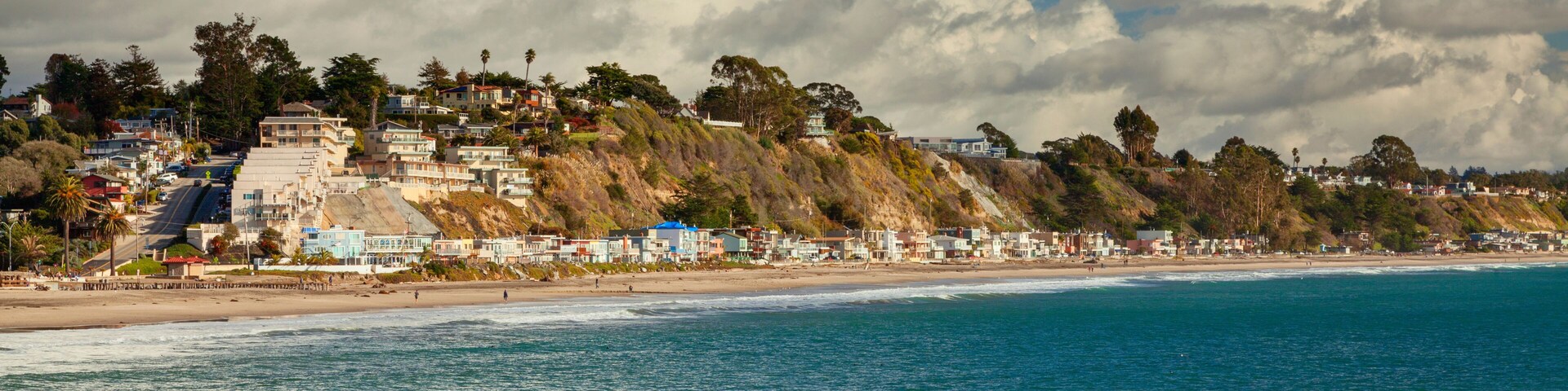 Uma vila costeira com uma praia, casas em uma encosta e uma grande nuvem no céu.