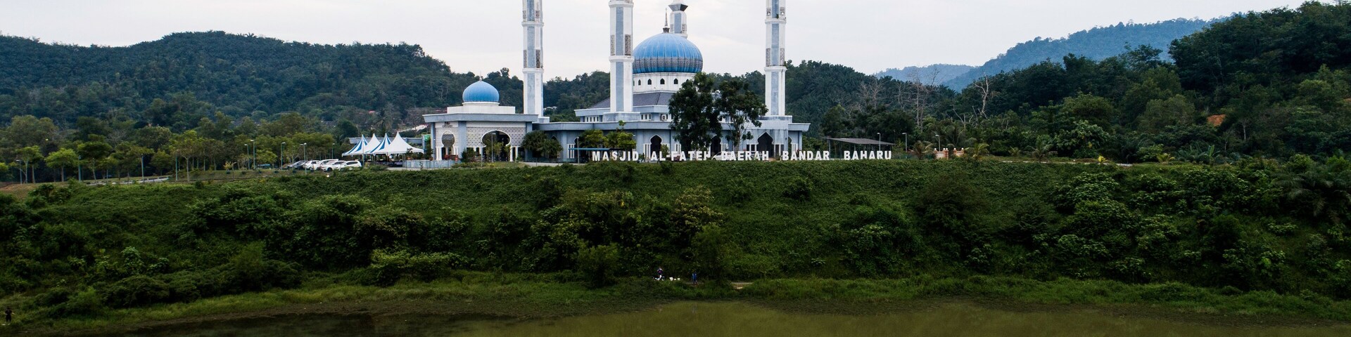 Uma mesquita com quatro minaretes e uma cúpula azul, rodeada por vegetação exuberante e um lago calmo.