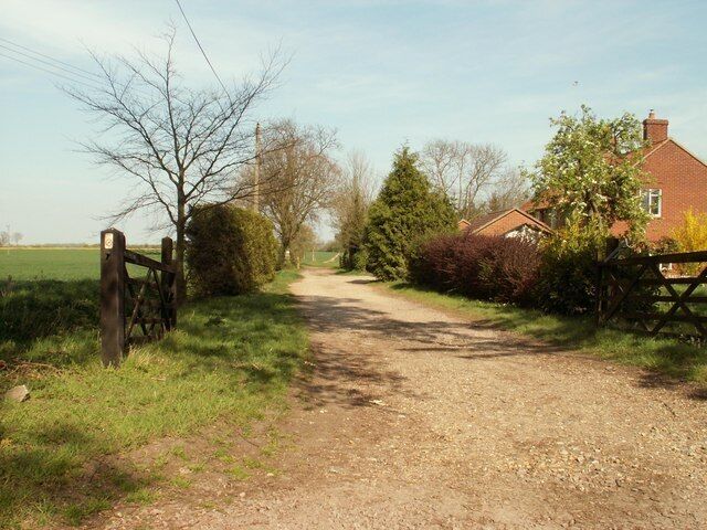 Uma estrada de terra rural que leva a uma propriedade murada, cercada por árvores e uma casa de tijolos.