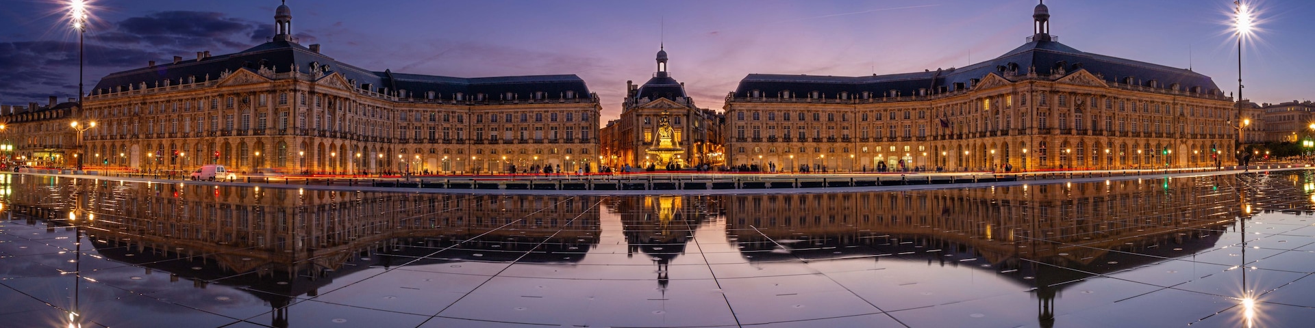 Um edifício imponente com uma cúpula central, acompanhado por duas alas simétricas, refletindo em uma superfície aquática calma.
