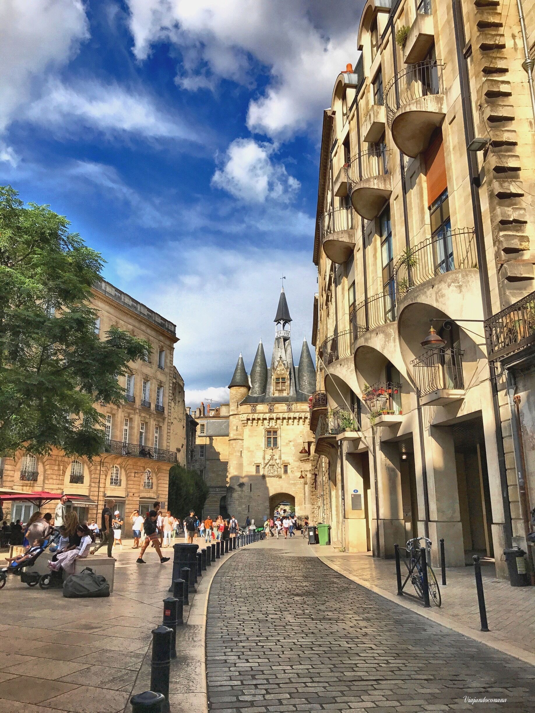 Uma rua de paraleipípedos flanqueada por edifícios históricos, uma torre imponente e um céu azul límpido.