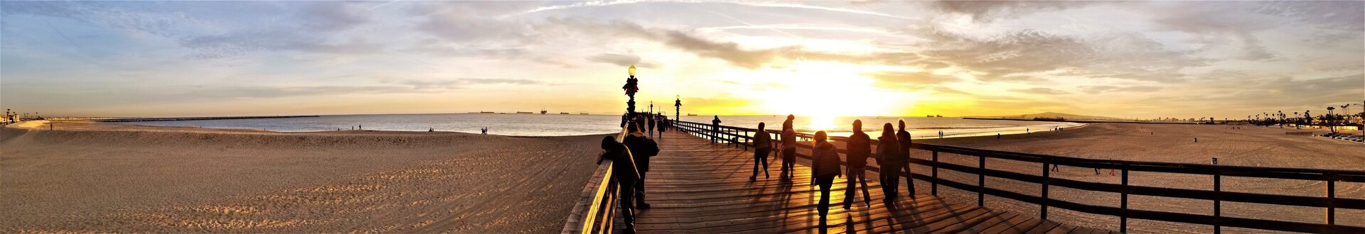 Uma praia com uma passarela de madeira que leva à água, um pôr do sol ao fundo e navios à distância.