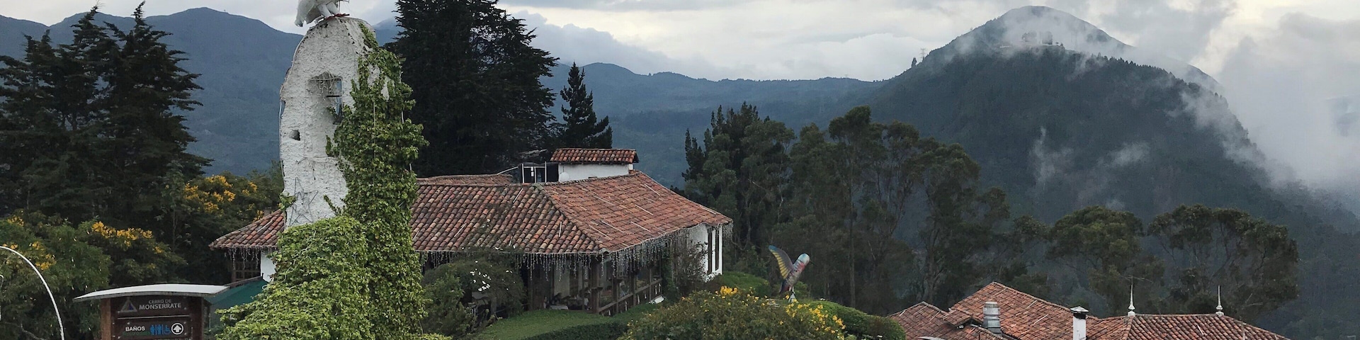 Paisagem montanhosa com uma estátua branca imponente, edifícios tradicionais com telhados de terracota e vegetação exuberante.