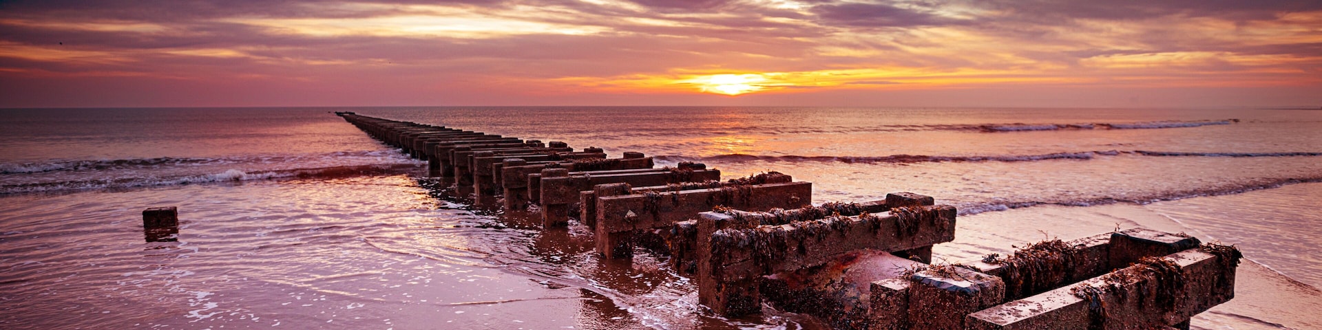 Um pôr do sol sobre uma praia com um píer enferrujado que se estende para a água.