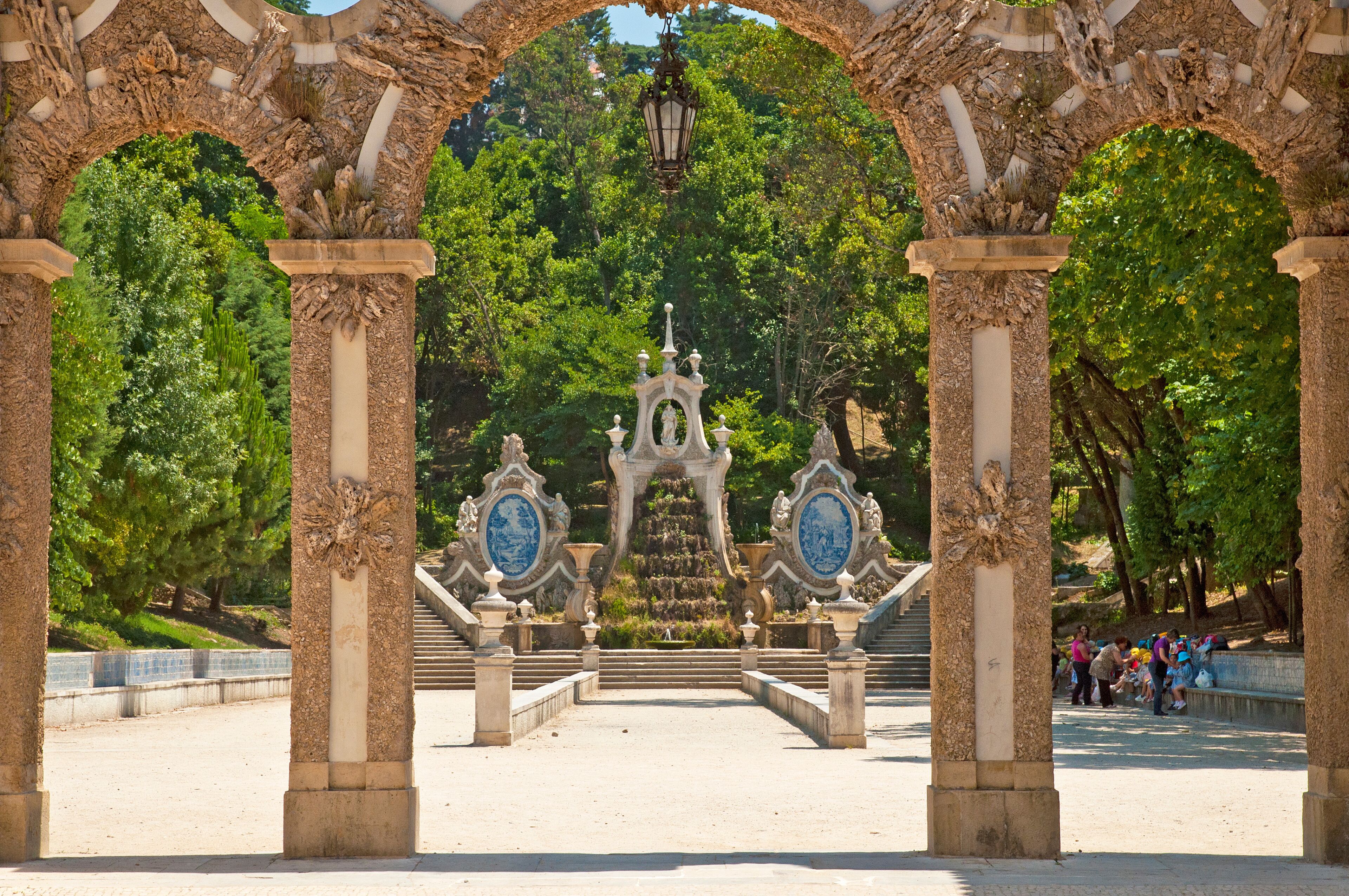 Uma entrada imponente com colunas ornamentadas e uma estátua central, cercada por vegetação exuberante.