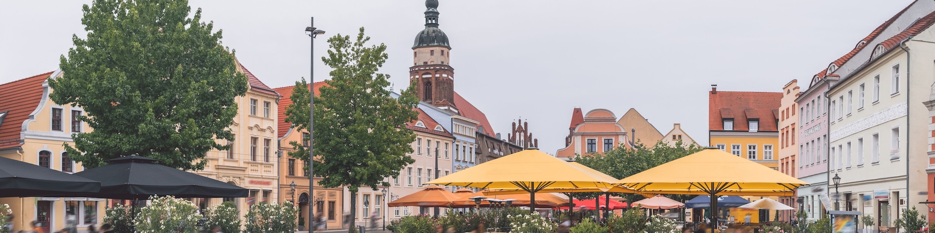 Uma praça movimentada com cafés ao ar livre, edifícios históricos e uma torre de igreja imponente.