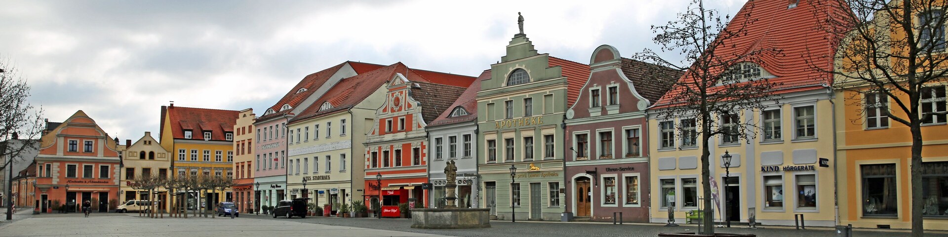 Uma praça histórica europeia com edifícios coloridos, uma estátua e um pavimento de pedra.